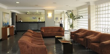Um hall de hotel com uma mesa de recepção, sofás marrons, uma mesa de centro e uma janela grande com painéis de vidro decorativos.
