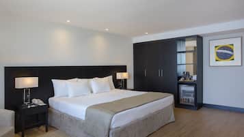 Quarto de hotel moderno com uma cama grande, mesas de cabeceira, minibar e televisão fixada na parede.