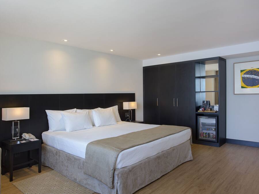 Quarto de hotel moderno com uma cama grande, mesas de cabeceira, minibar e televisão fixada na parede.
