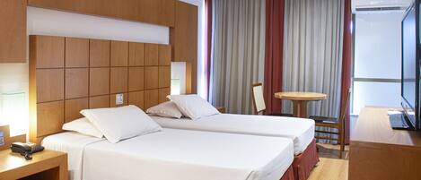 Quarto de hotel com duas camas, uma escrivaninha, uma cadeira, uma mesinha e uma janela com cortinas.