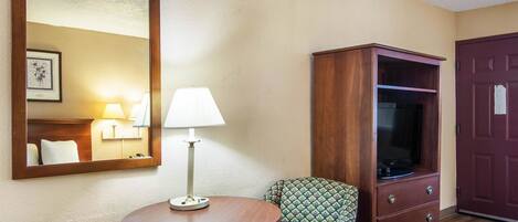 Quarto de hotel com uma mesa redonda, duas poltronas estampadas, um espelho grande, um armário de madeira com televisão e uma porta.