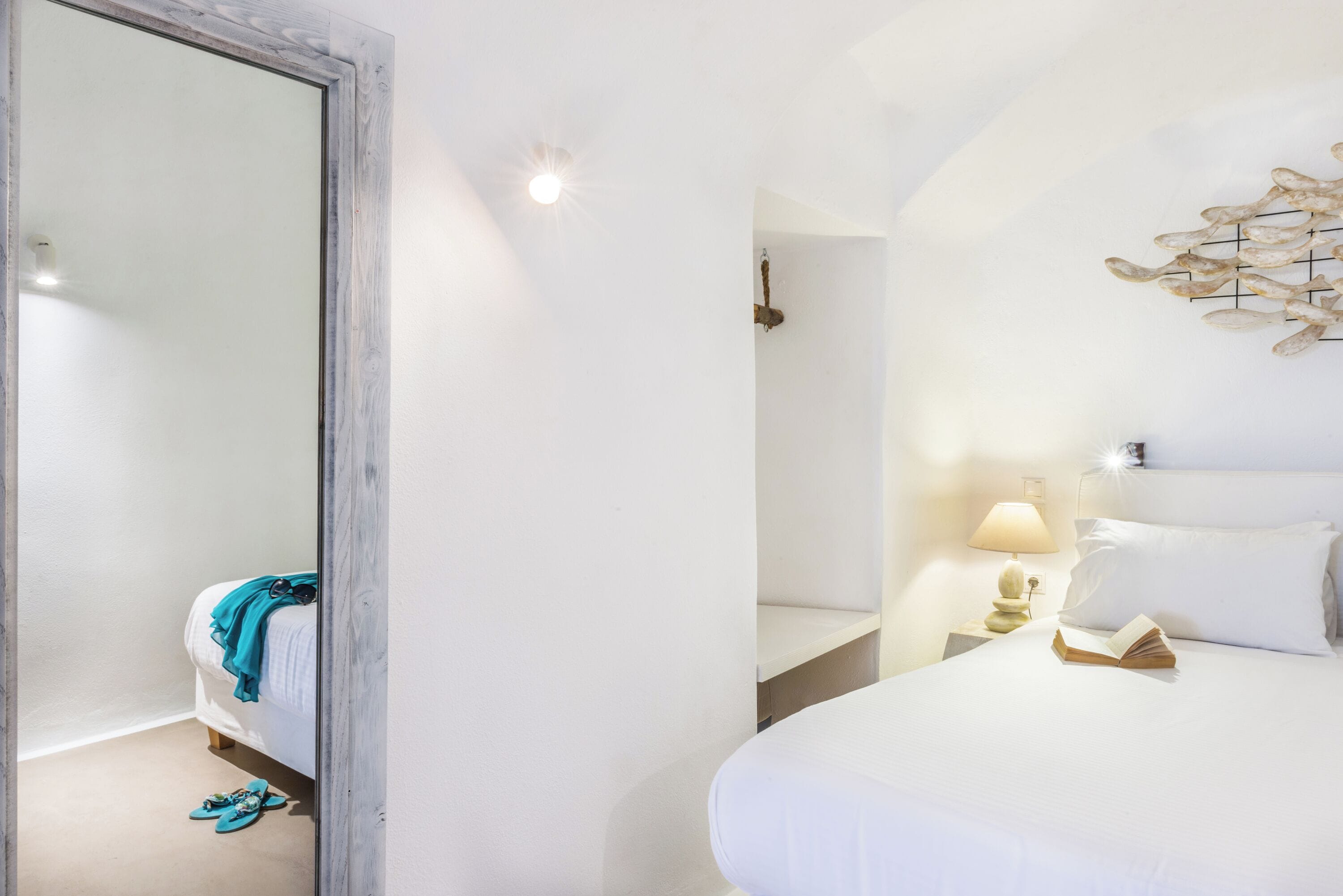 Quarto de hotel com um espelho grande, uma cama com roupa de cama branca, uma mesa de cabeceira com abajur e um candeeiro fixo na parede.