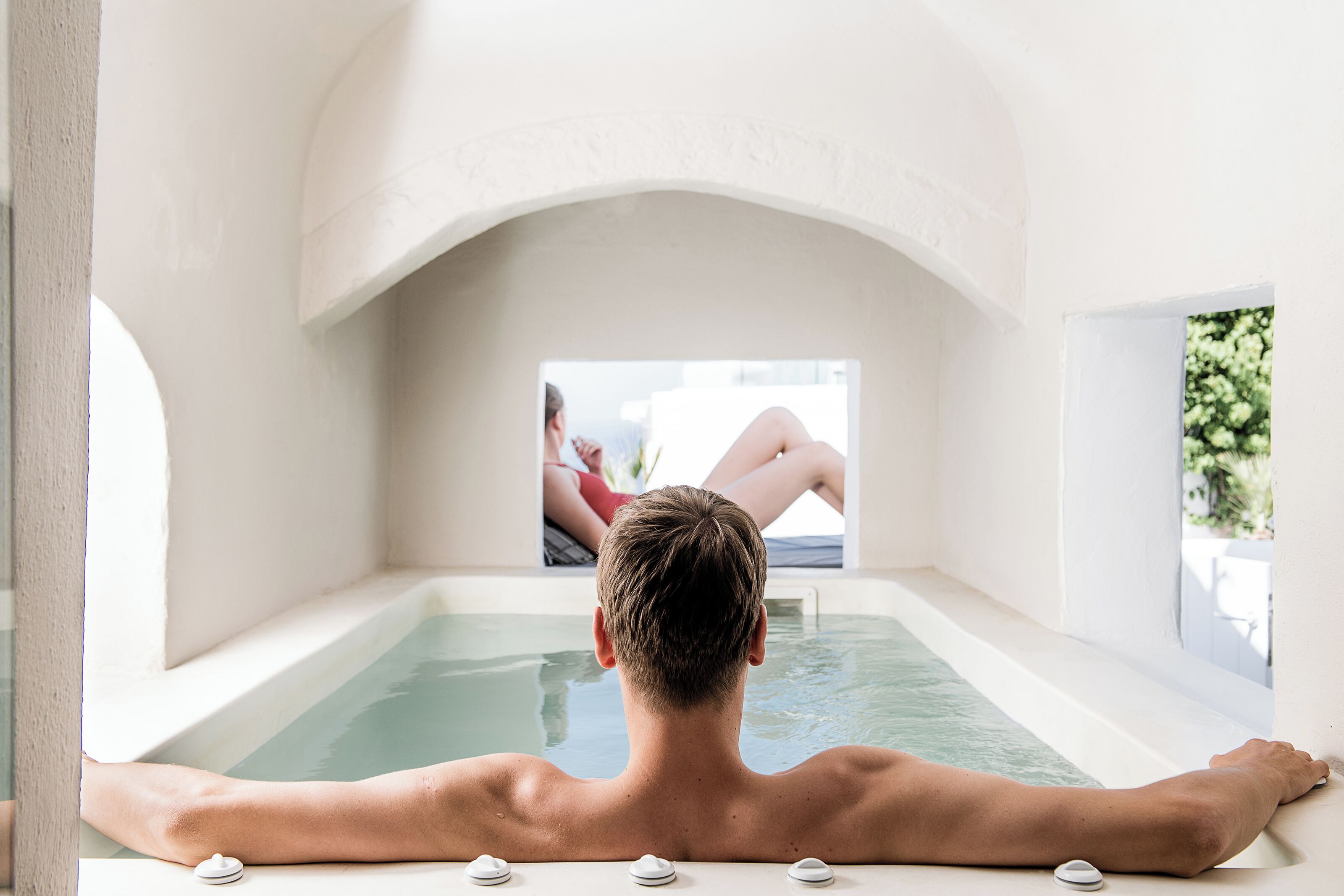 Um homem relaxa em uma piscina infinita branca, com vista para uma mulher de maiô vermelho.