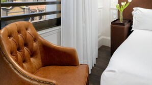 Quarto de hotel com vista para uma catedral, uma poltrona de couro marrom, uma mesinha de cabeceira com um vaso de flores e um candeeiro pendente.