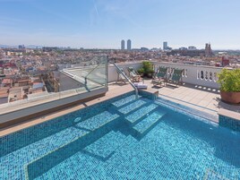 Piscina no terraço com guarda-corpo de vidro, cadeiras de descanso e vista para a cidade.