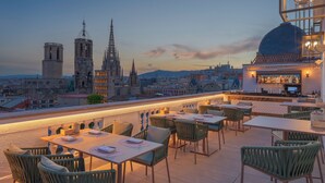 Um bar no terraço com mesas e cadeiras, uma paisagem urbana ao fundo e um espaço de bar com balcão e iluminação.