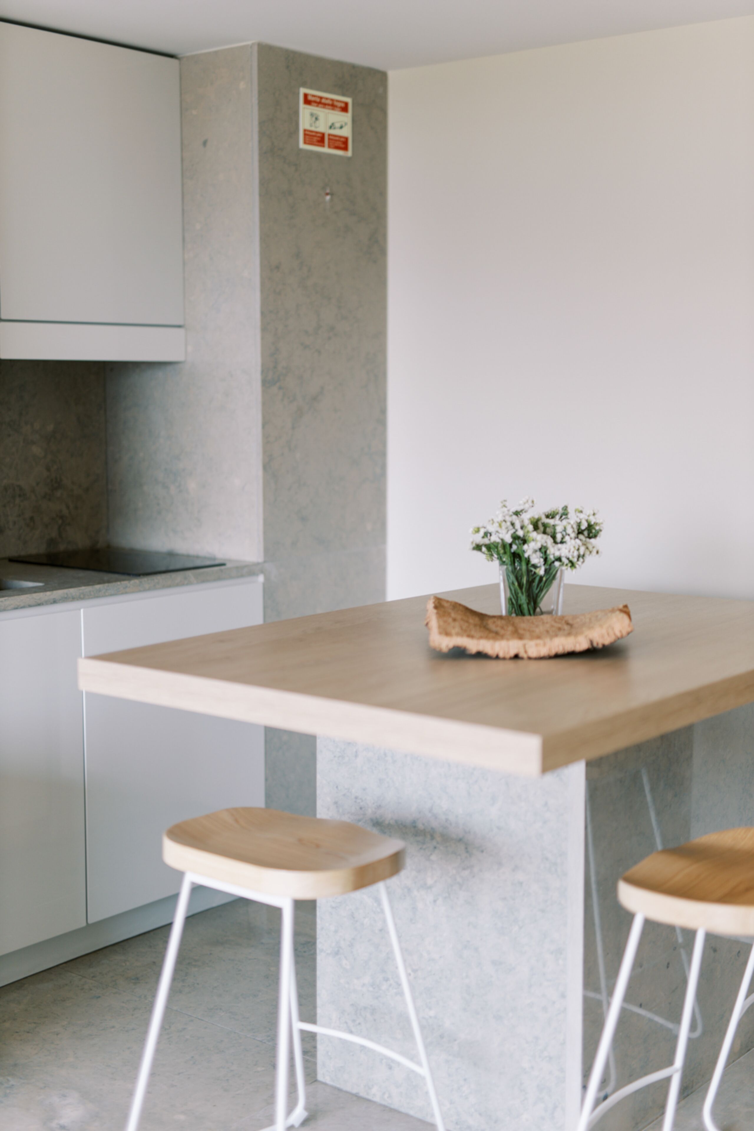 Uma cozinha moderna com mesa e banquetas de madeira, um vaso com flores e um aviso na parede.