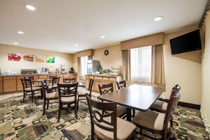 Uma área de refeições de hotel com uma grande mesa de madeira e cadeiras, uma cozinha com máquina de café e uma TV de tela plana presa na parede.