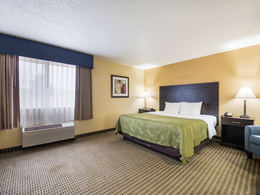 Quarto de hotel com cama, duas mesas de cabeceira com luminárias, uma janela com cortinas e ar condicionado fixado na parede.
