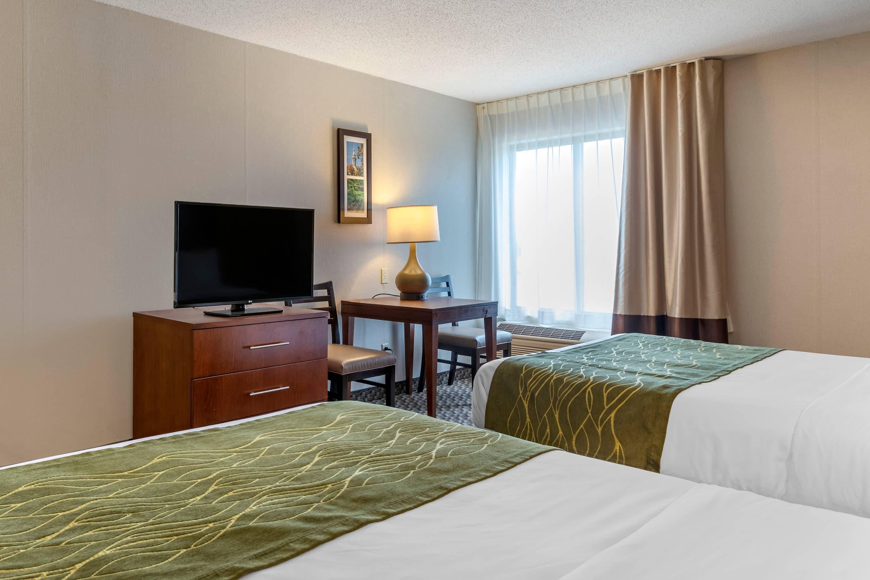 Quarto de hotel com duas camas, uma mesa de trabalho, televisão, luminária e uma cadeira.