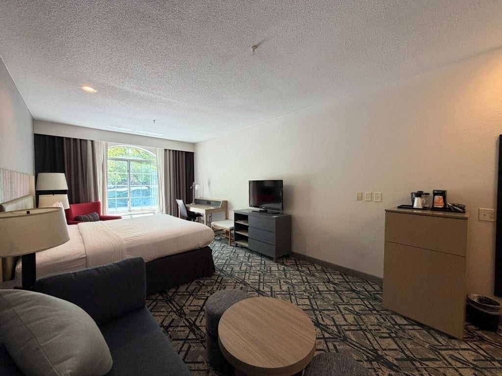 Quarto de hotel com cama, sofá, televisão, escrivaninha e uma mesinha redonda.
