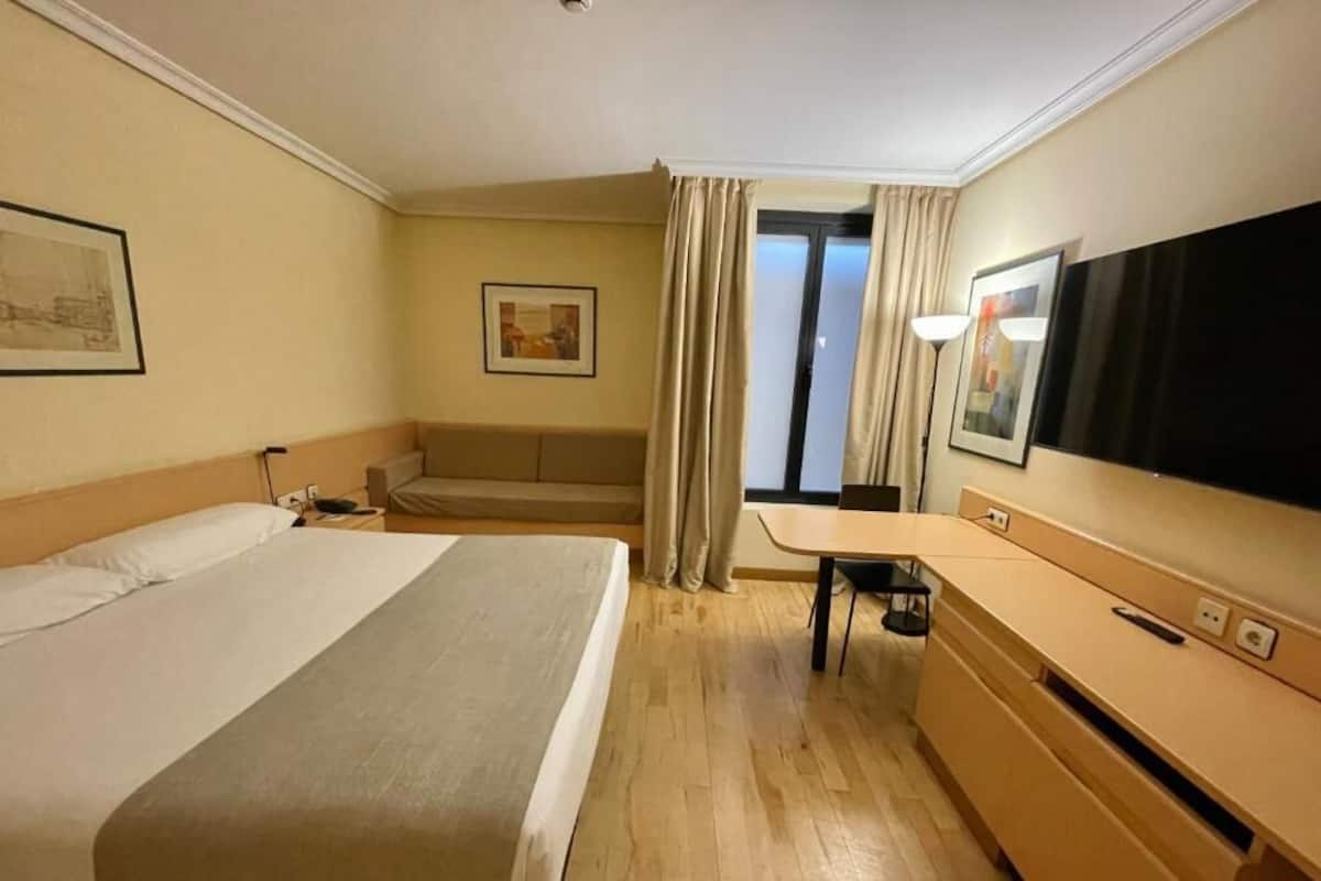 Quarto de hotel com cama, escrivaninha, cadeira, televisão e janela com cortinas.