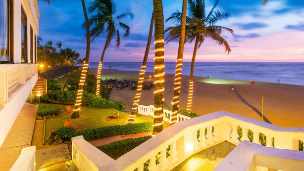 Hotel à beira-mar com palmeiras iluminadas, piscina e vista para o oceano.