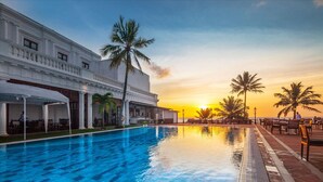 Piscina com vista para o pôr do sol, palmeiras e um edifício com colunas.