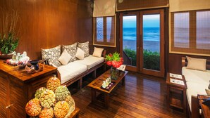 Quarto aconchegante com piso de madeira, sofá com almofadas estampadas, mesa de jantar com um vaso de flores e vista para o oceano por meio de grandes portas deslizantes.
