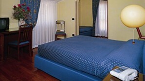Quarto de hotel com cama, escrivaninha, cadeira, televisão e luminária.