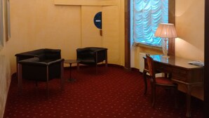 Quarto de hotel com tapete vermelho, escrivaninha com luminária, duas cadeiras, janela grande com cortinas azuis e um quadro na parede.