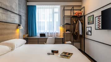 Quarto de hotel com cama, escrivaninha com laptop, cadeira, guarda-roupa e janela com cortinas.