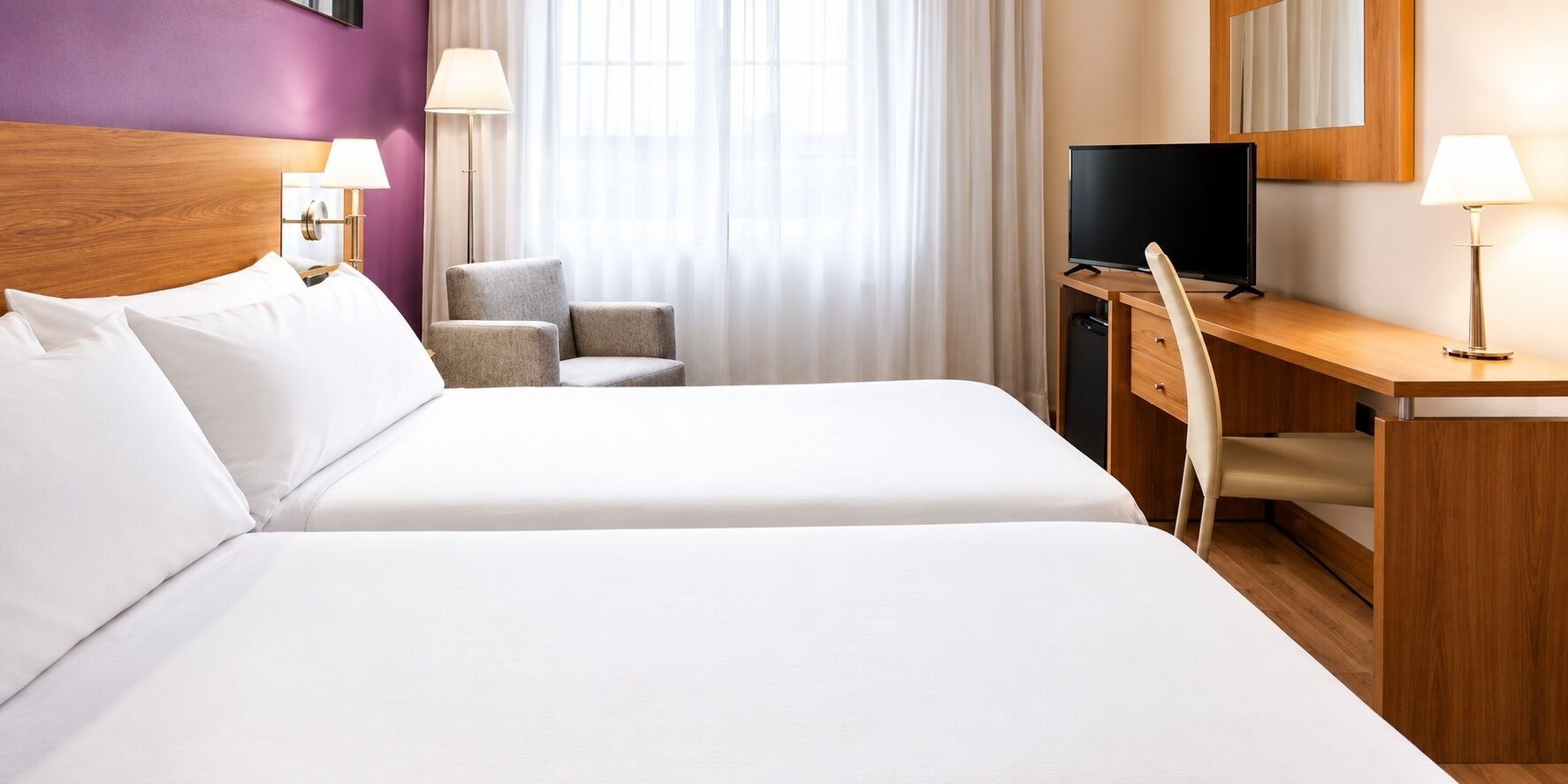 Quarto de hotel com duas camas, uma escrivaninha, televisão, uma cadeira e um abajur.