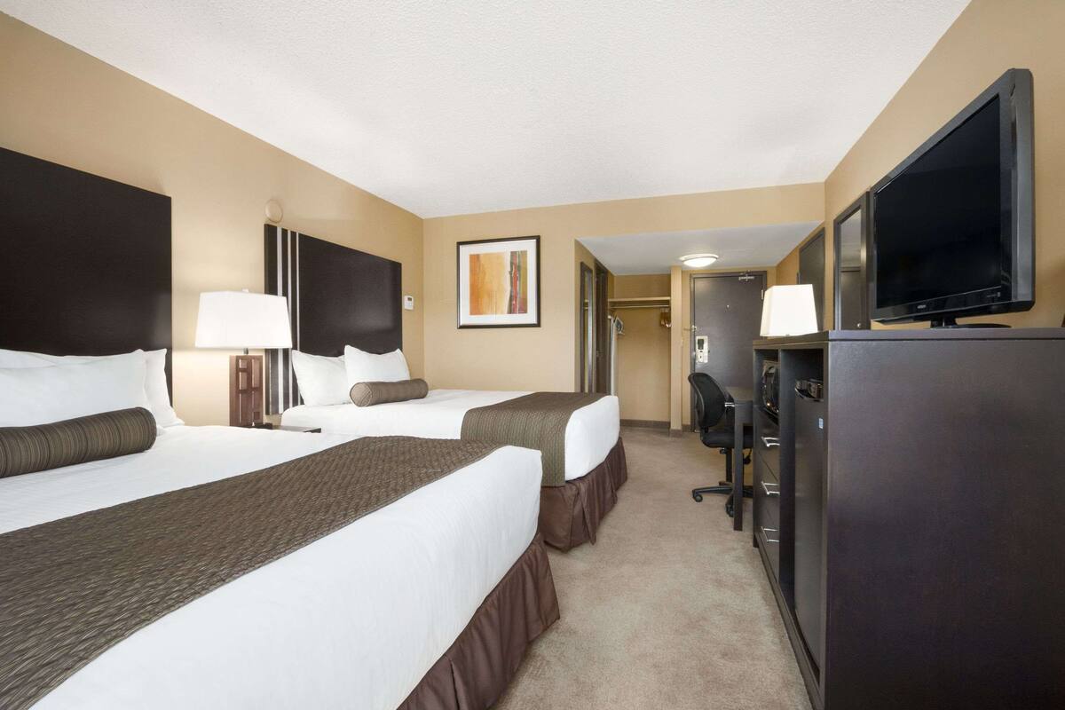Quarto de hotel com duas camas, uma escrivaninha, televisão e banheiro visível através de uma porta aberta.