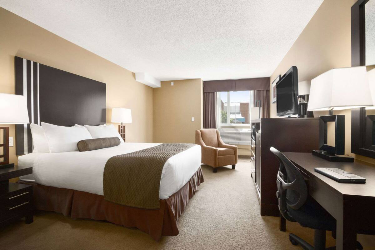 Quarto de hotel com cama, escrivaninha, cadeira, televisão e janela com cortinas.