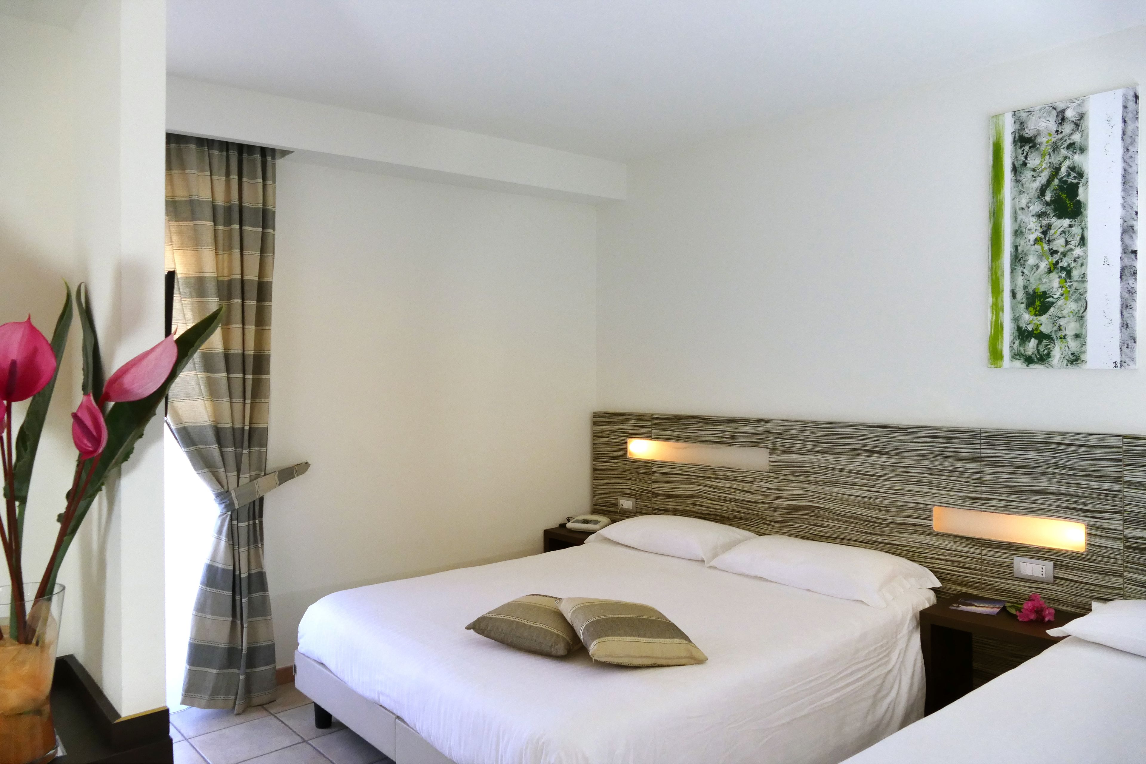 Quarto de hotel com cama, mesas de cabeceira, um vaso com flores e cortina.