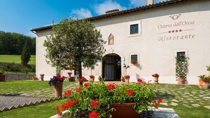 Um edifício com uma placa que diz 'Osteria dell'Orcia Ristorante', rodeado por um jardim com plantas e flores em vasos.
