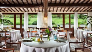 Restaurante com mesas redondas, toalhas brancas e cadeiras de madeira. Grandes janelas proporcionam vista para uma paisagem verde.