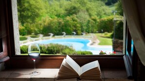 Janela com um livro aberto e um copo de vinho tinto, com vista para uma piscina e vegetação exuberante.