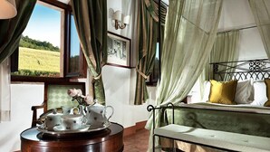 Quarto com cama, mesa de cabeceira com bandeja de chá, quadro na parede e vista para um campo verde pela janela.