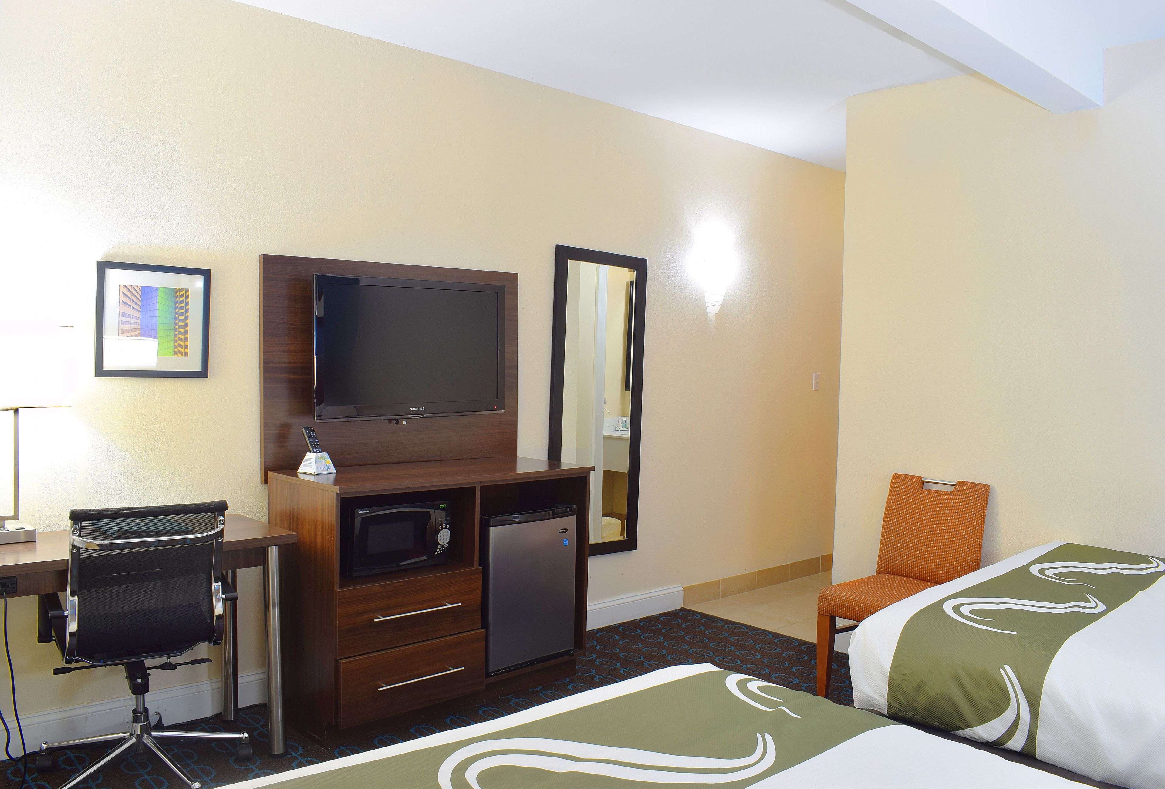 Quarto de hotel com escrivaninha, televisão, micro-ondas, frigorífico, cama e cadeira.