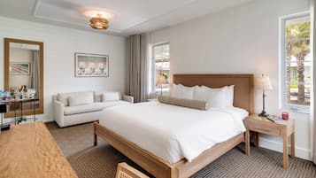Quarto de hotel com uma cama grande, uma mesinha de cabeceira de madeira, um criado-mudo, uma escrivaninha, uma cadeira, um sofá, uma janela ampla e um espelho.