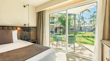 Quarto de hotel com cama, mesa de cabeceira, luminária e vista para área da piscina com cadeiras e palmeiras.