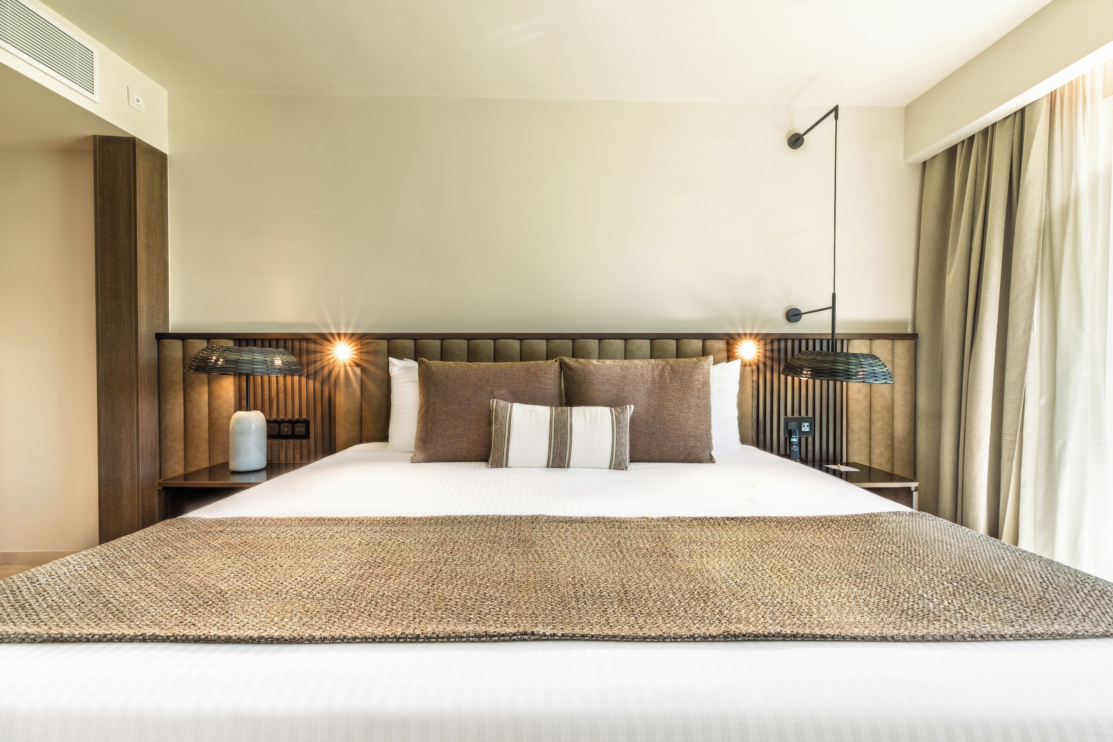 Quarto de hotel com uma cama grande, cabeceira com iluminação integrada, mesa de cabeceira com abajur e vista para o exterior através de uma janela com cortinas.