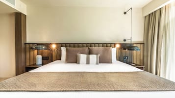 Quarto de hotel com uma cama grande, cabeceira com iluminação integrada, mesa de cabeceira com abajur e vista para o exterior através de uma janela com cortinas.