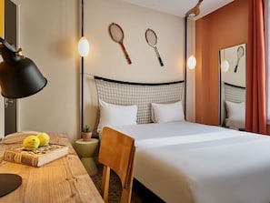 Quarto de hotel com cama, escrivaninha com abajur, cadeira, mesinha com um livro e bolas de ténis, e duas raquetes de ténis na parede.