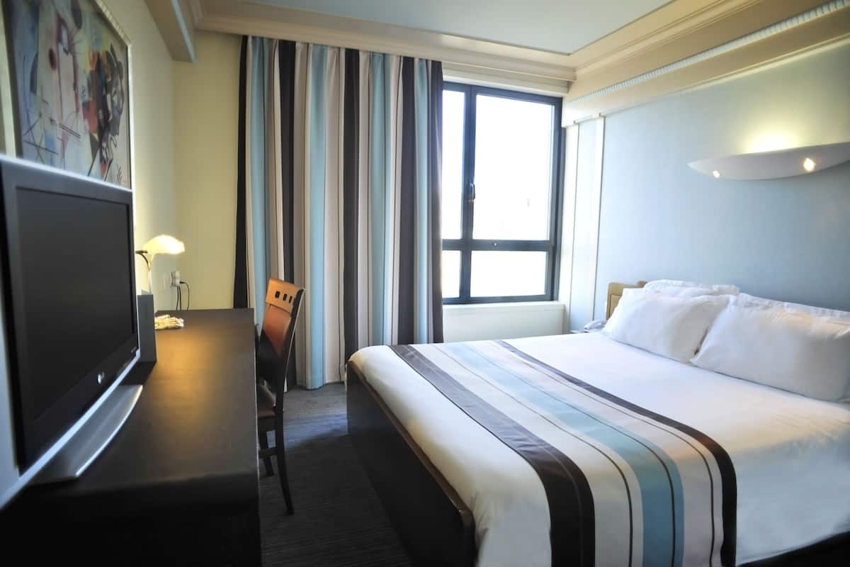 Quarto de hotel com cama, secretária com televisão, cadeira e janela com cortinas.