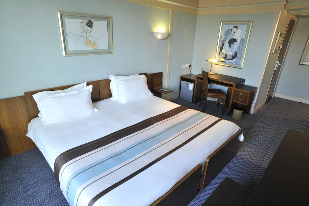 Quarto de hotel com uma cama grande, uma escrivaninha e uma cadeira. Há duas imagens emolduradas na parede.