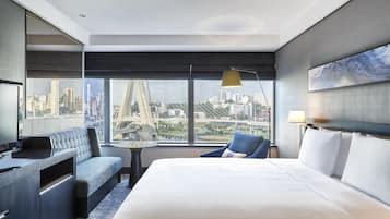 Quarto de hotel com cama, escrivaninha, cadeira e vista para a cidade através de grandes janelas.