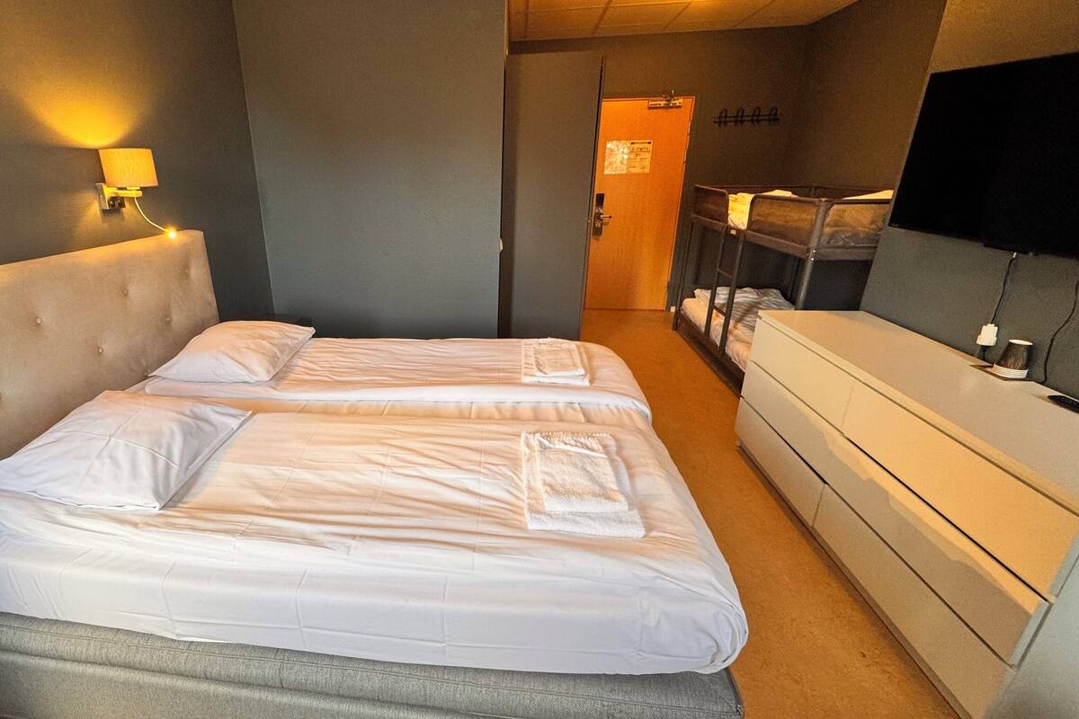 Quarto de hotel com cama de casal, luminárias de cabeceira, televisão, cômoda e porta que dá para outro ambiente.
