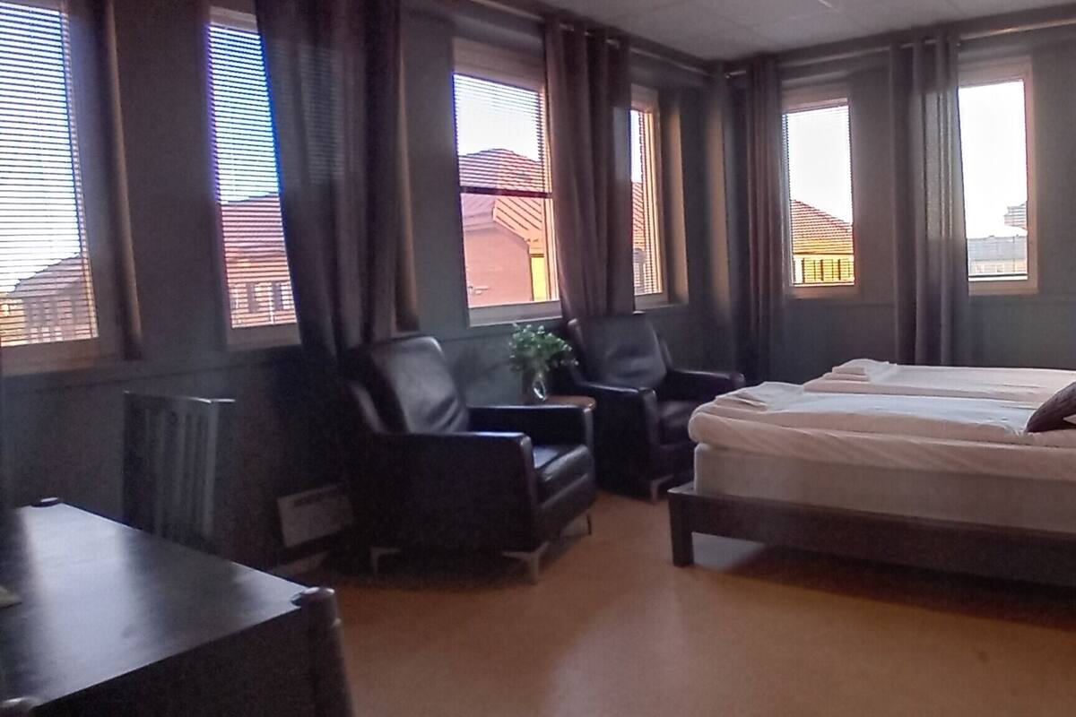 Quarto de hotel com uma cama, duas cadeiras, uma mesa de trabalho e janelas amplas.