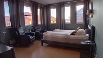 Quarto de hotel com uma cama, duas cadeiras, uma mesa de trabalho e janelas amplas.