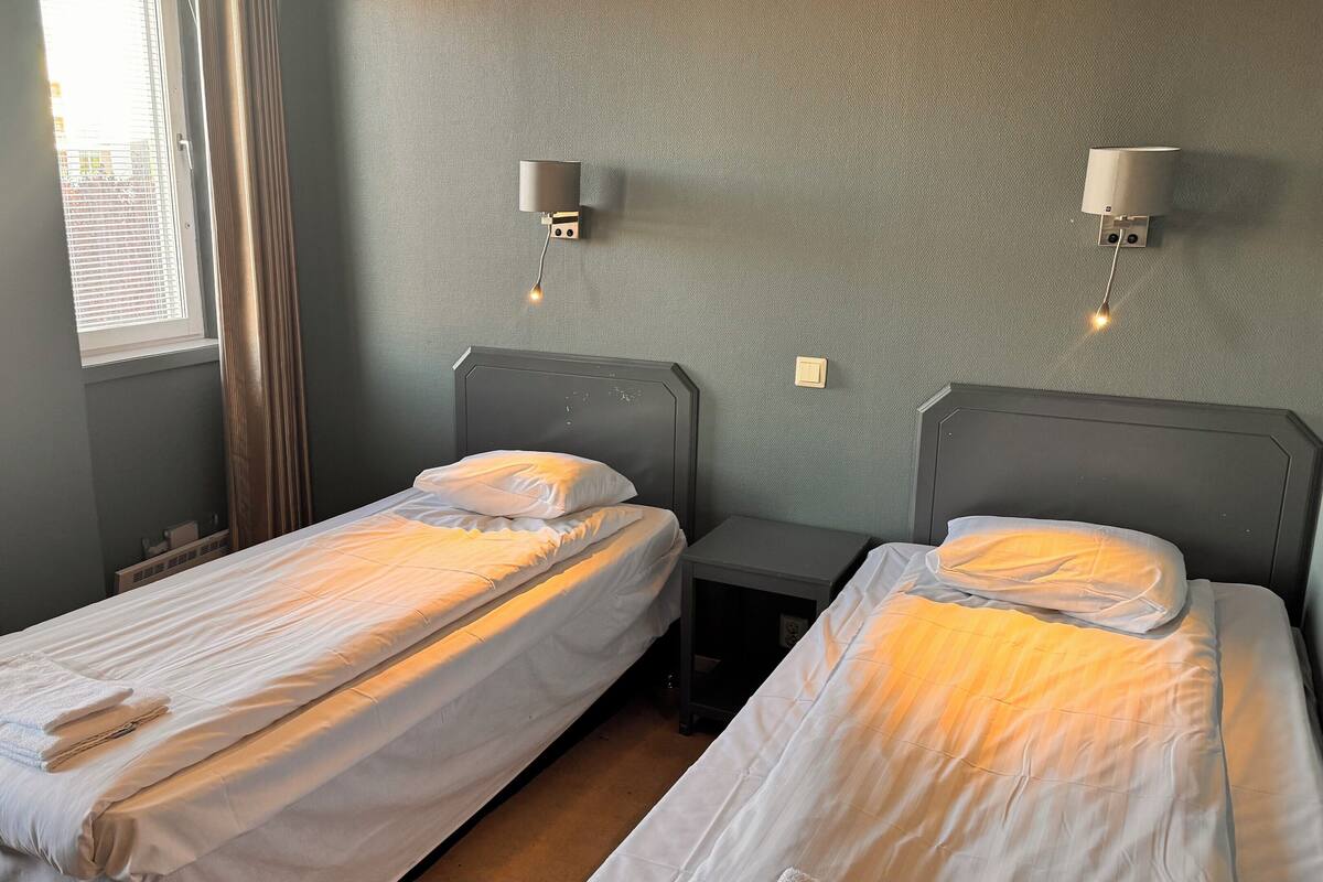 Quarto de hotel com duas camas de solteiro, mesa de cabeceira, luminária na parede e janela com cortinas.