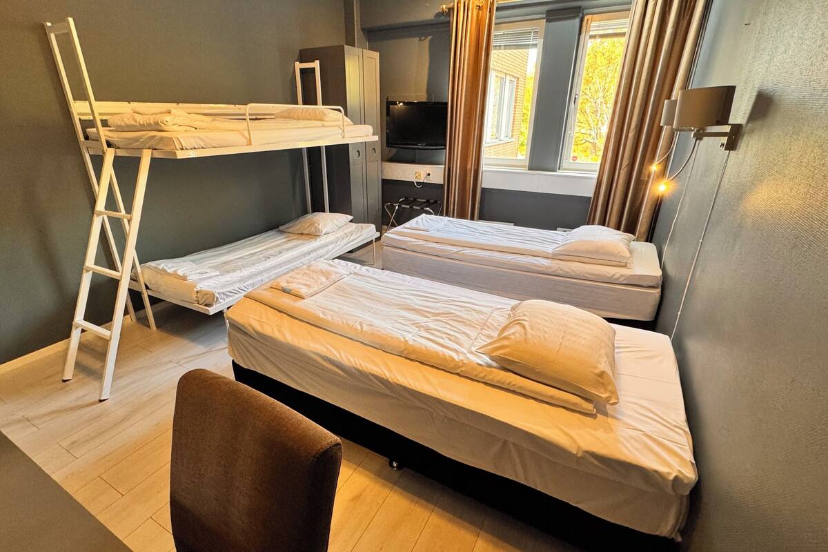 Quarto com beliches, escrivaninha, cadeira, televisão e janela com cortinas.