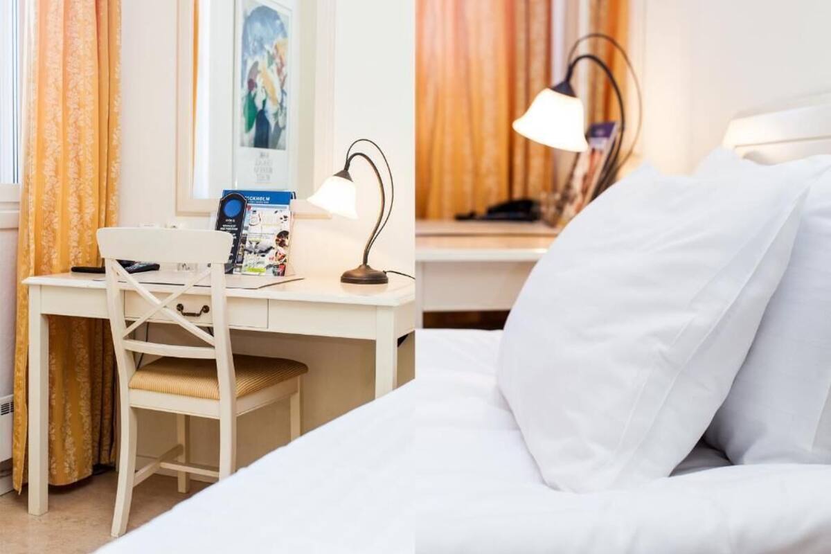 Quarto de hotel com uma escrivaninha branca, uma cadeira, uma luminária e uma cama com roupa de cama branca.