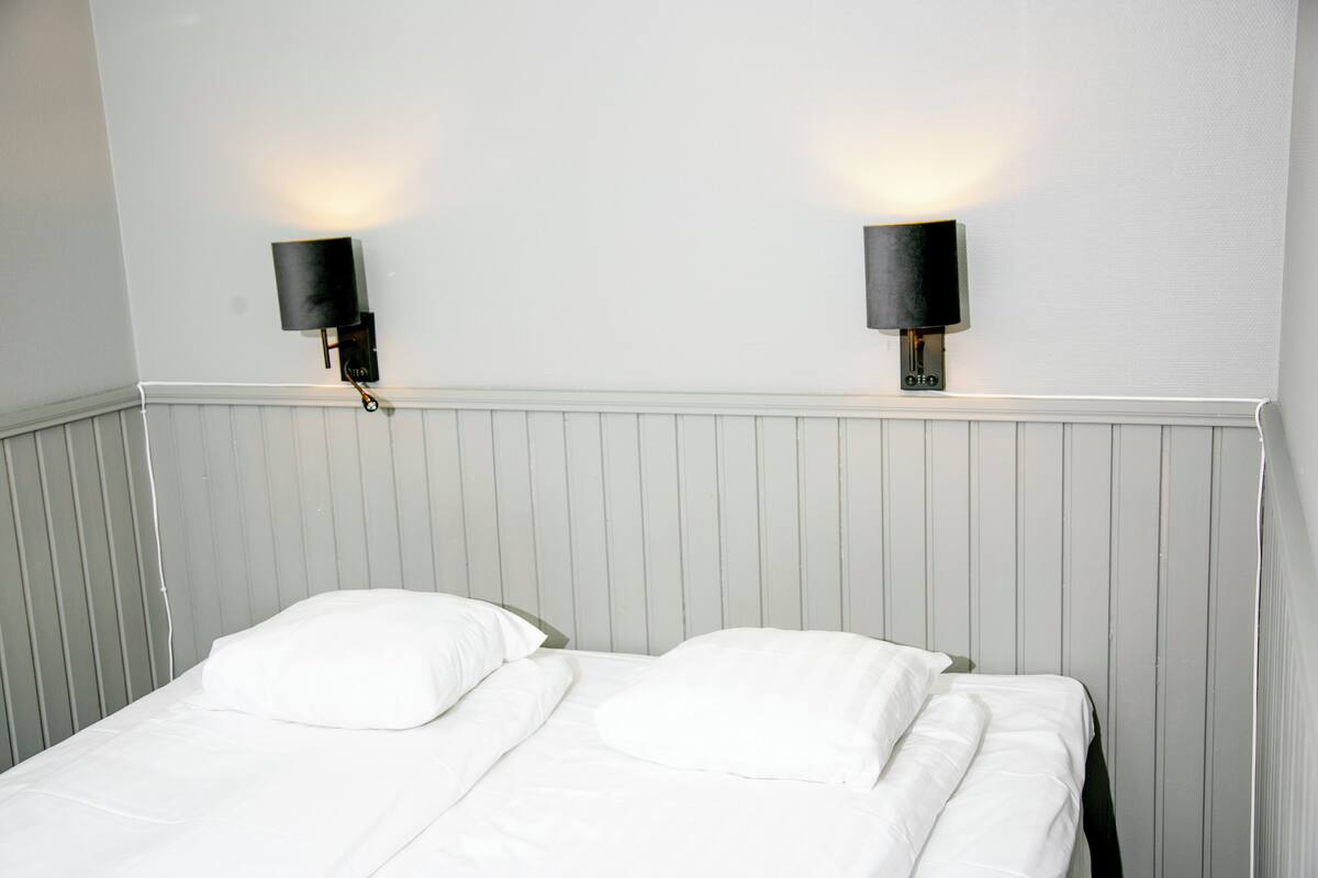 Cama bem arrumada com lençóis brancos e duas luminárias fixadas na parede, uma de cada lado.