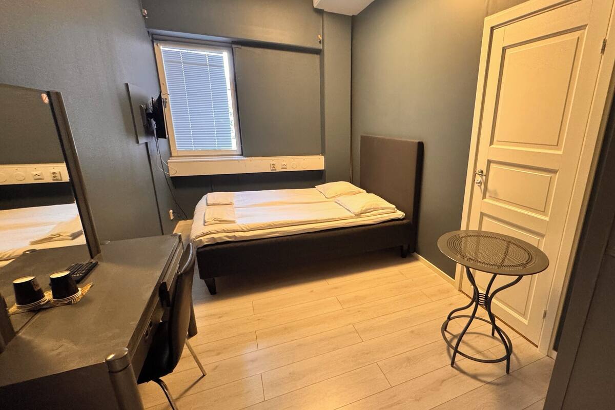 Quarto compacto com uma cama de solteiro, uma mesinha, uma escrivaninha com um computador e uma janela com persianas.