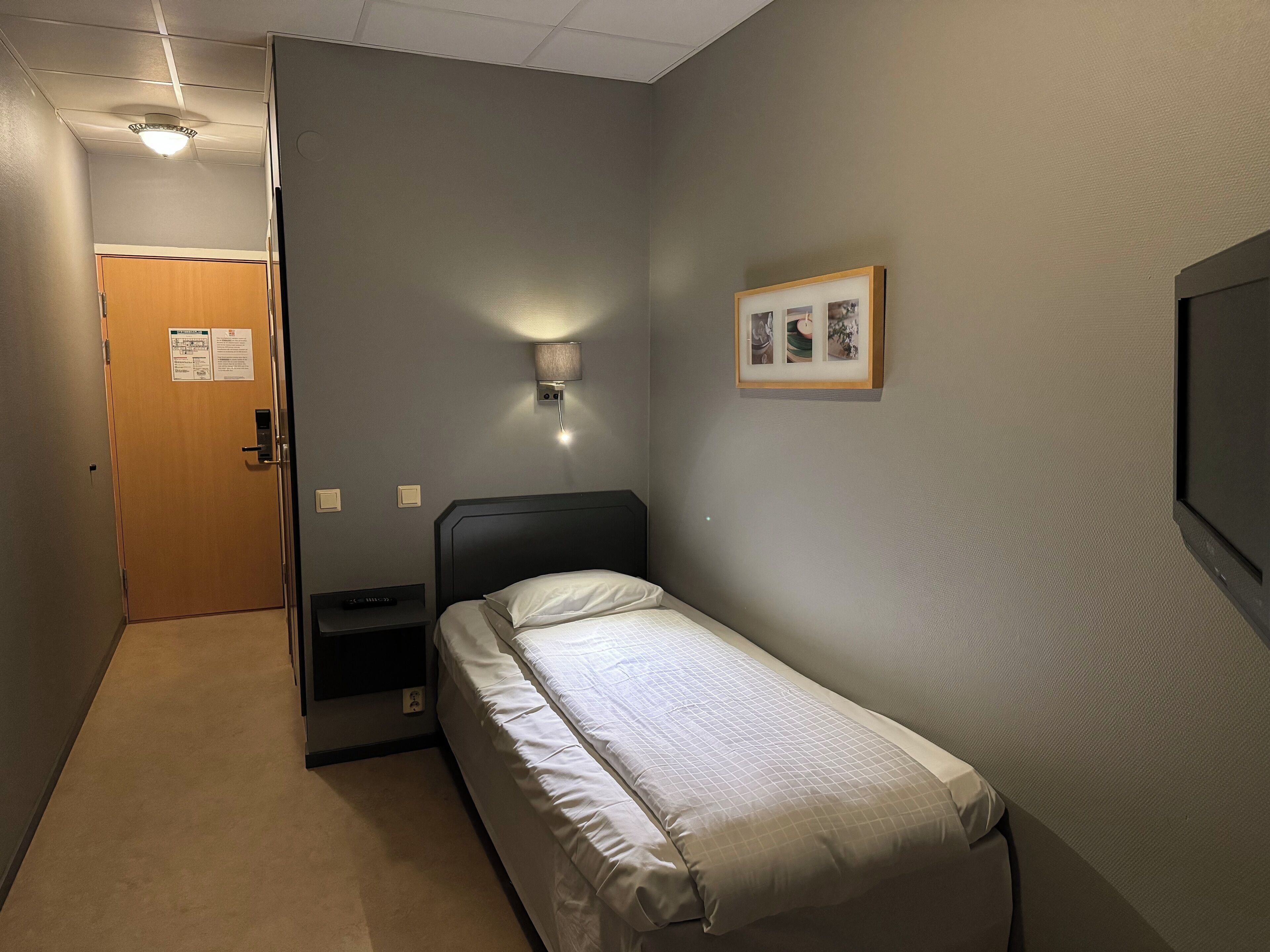 Quarto pequeno com uma cama de solteiro, porta de madeira, mesa de cabeceira, luz acoplada na parede e um quadro enquadrado.