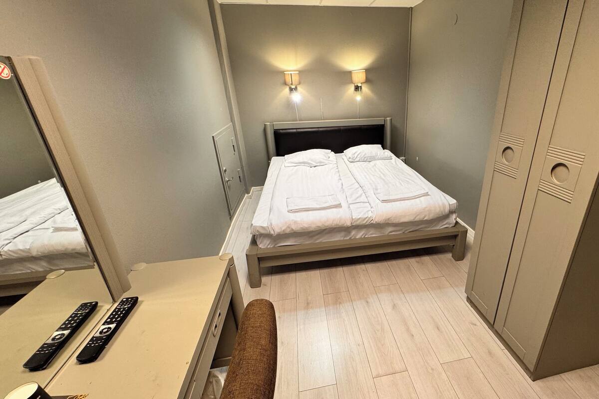Quarto compacto com cama, mesa de cabeceira, espelho e guarda-roupa.