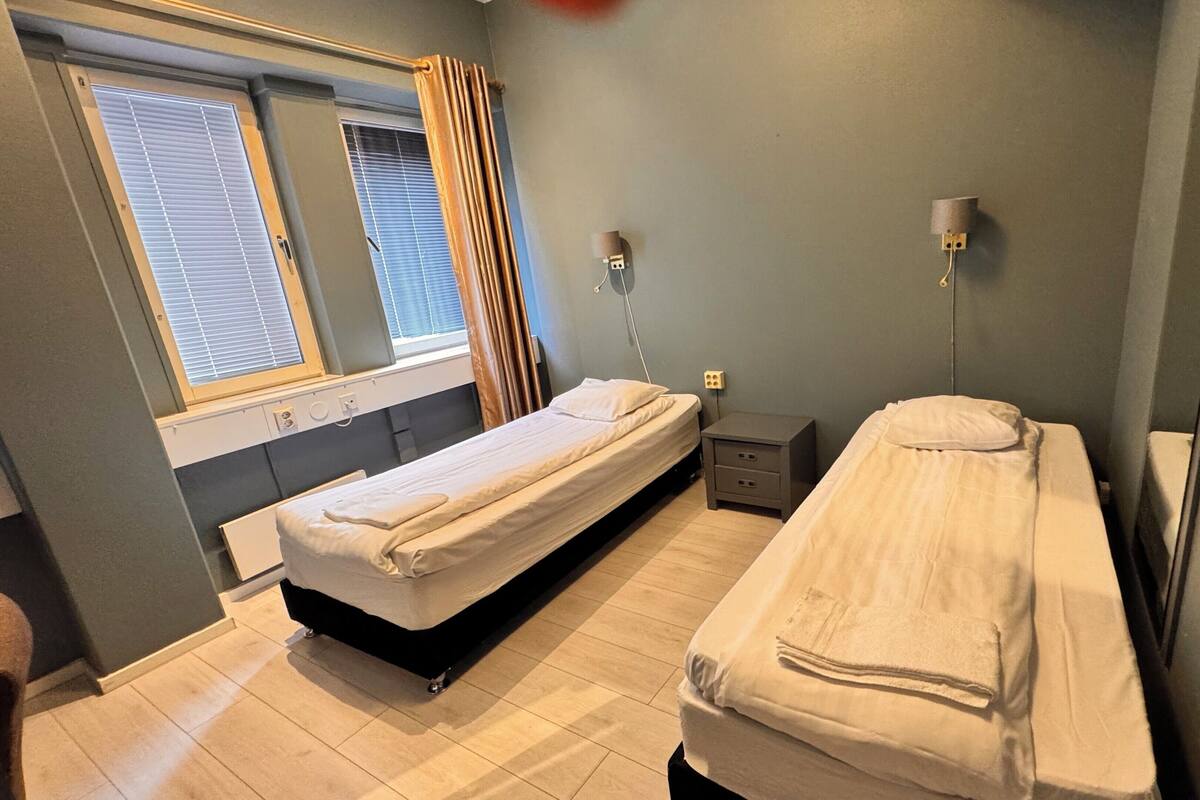 Quarto de hotel pequeno e individual com duas camas de solteiro, janela com persianas, mesa de cabeceira e luminária fixada na parede.